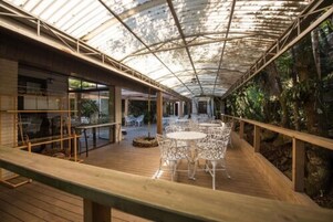 Espaço interno com deck de madeira, mesas e cadeiras de metal branco, cercado por paredes de vidro e estrutura de telhado metálico.