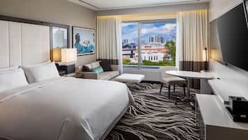 Quarto de hotel moderno com uma cama grande, uma escrivaninha, uma cadeira, uma mesinha e uma janela ampla com vista para a cidade.