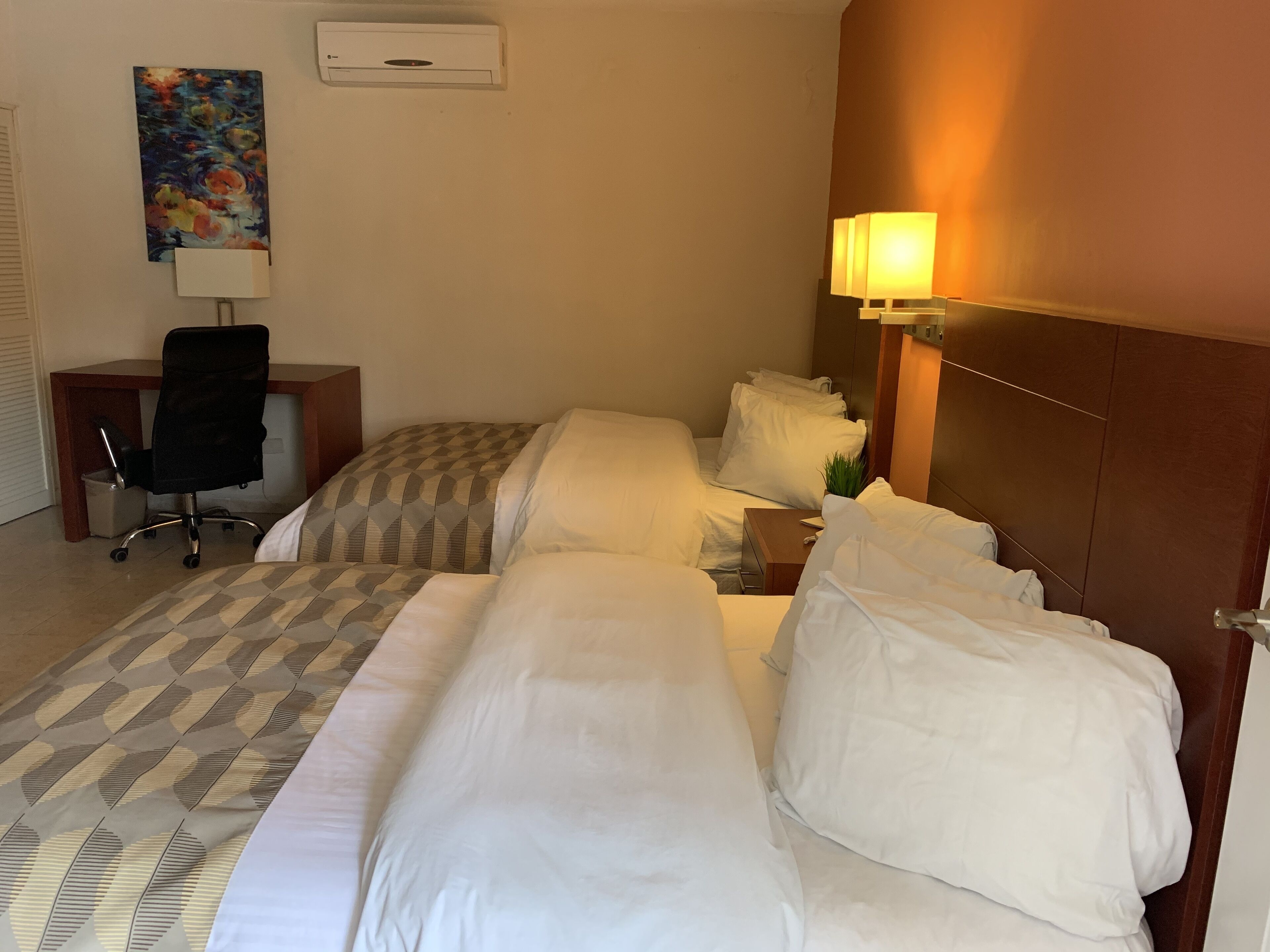 Quarto de hotel com duas camas, uma escrivaninha, uma cadeira, um abajur e um quadro na parede.