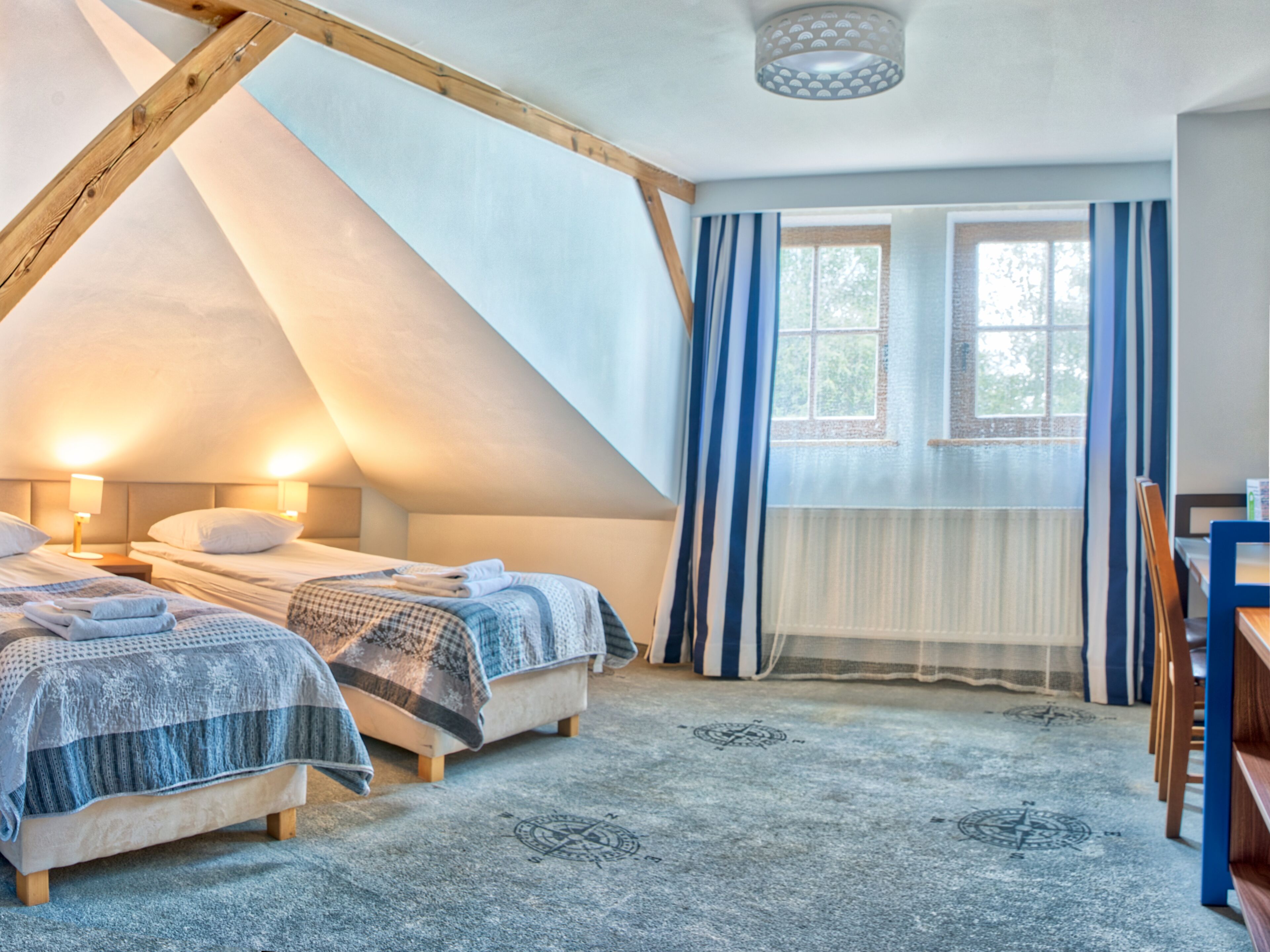Quarto aconchegante com duas camas, janela com cortinas, teto de madeira e tapete com desenho de bússola.