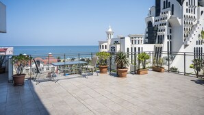 Terraço no telhado com plantas em vasos, uma mesa e cadeiras, com vista para uma paisagem urbana costeira.