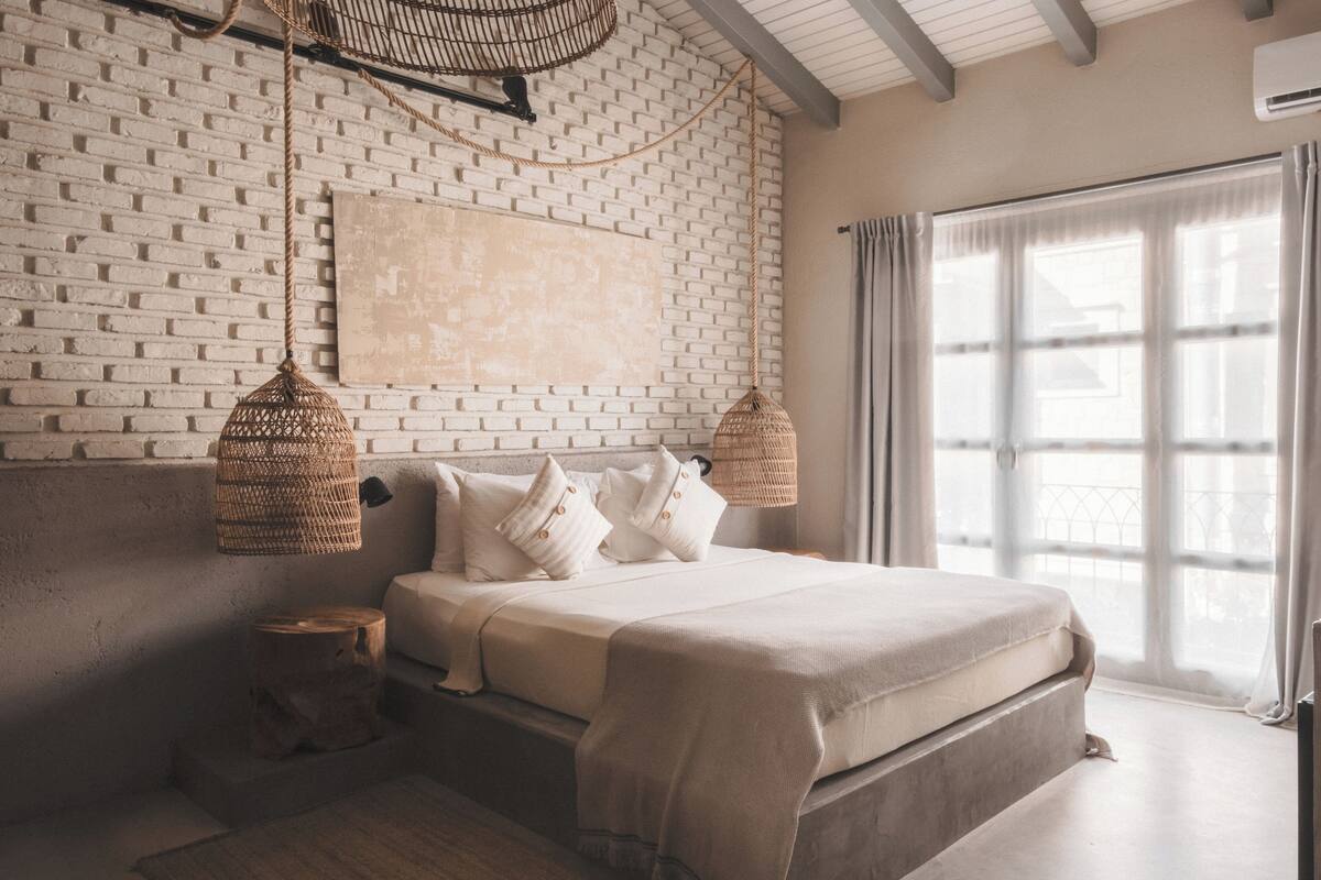 Quarto com parede de tijolos, uma cama grande com colcha bege, duas luminárias de vime suspensas e uma janela com cortinas.