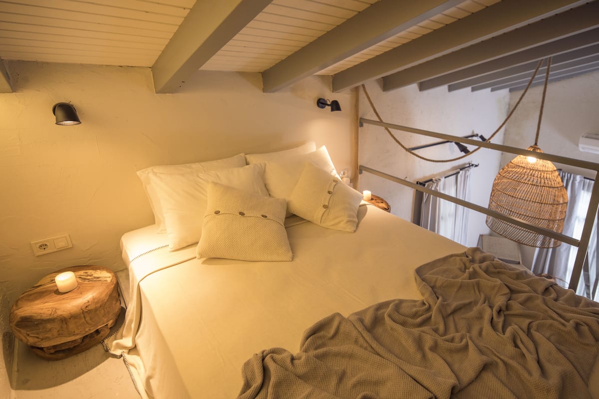 Cama bem arrumada com travesseiros e coberta, luminária de cabeceira, e mesa de cabeceira rústica de madeira com uma luminária.