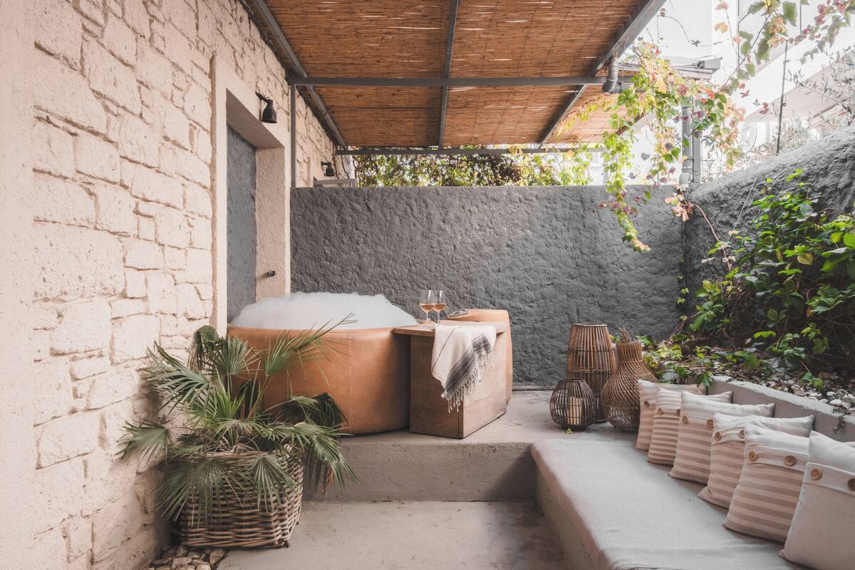 Uma área de estar aconchegante ao ar livre, com uma parede de pedra, pérgola de madeira e diversos almofados e plantas.