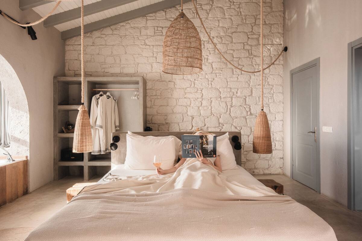 Um quarto aconchegante com parede de pedra, guarda-roupa de madeira, luminária de vime pendente e mesa de cabeceira com livro e copo.
