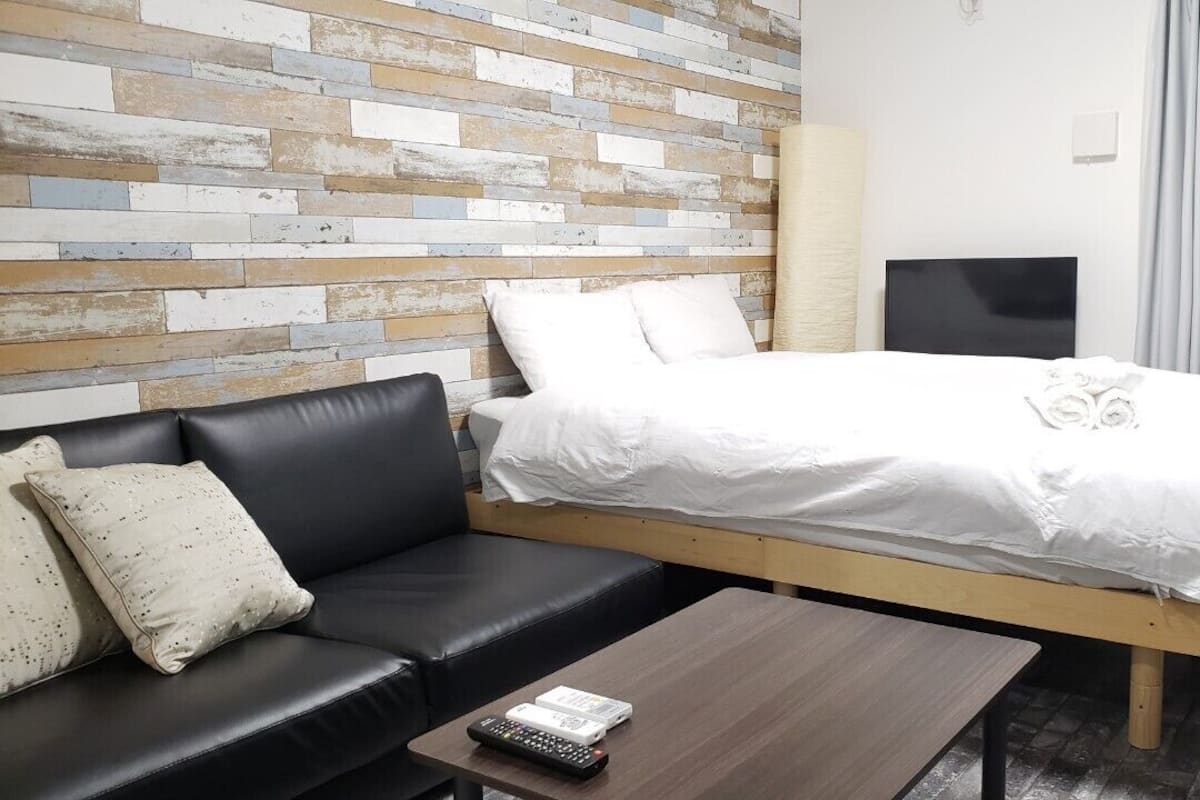 Quarto de hotel moderno com cama de cabeceira de madeira, sofá de couro preto, pequena mesa de madeira e televisão de tela plana.