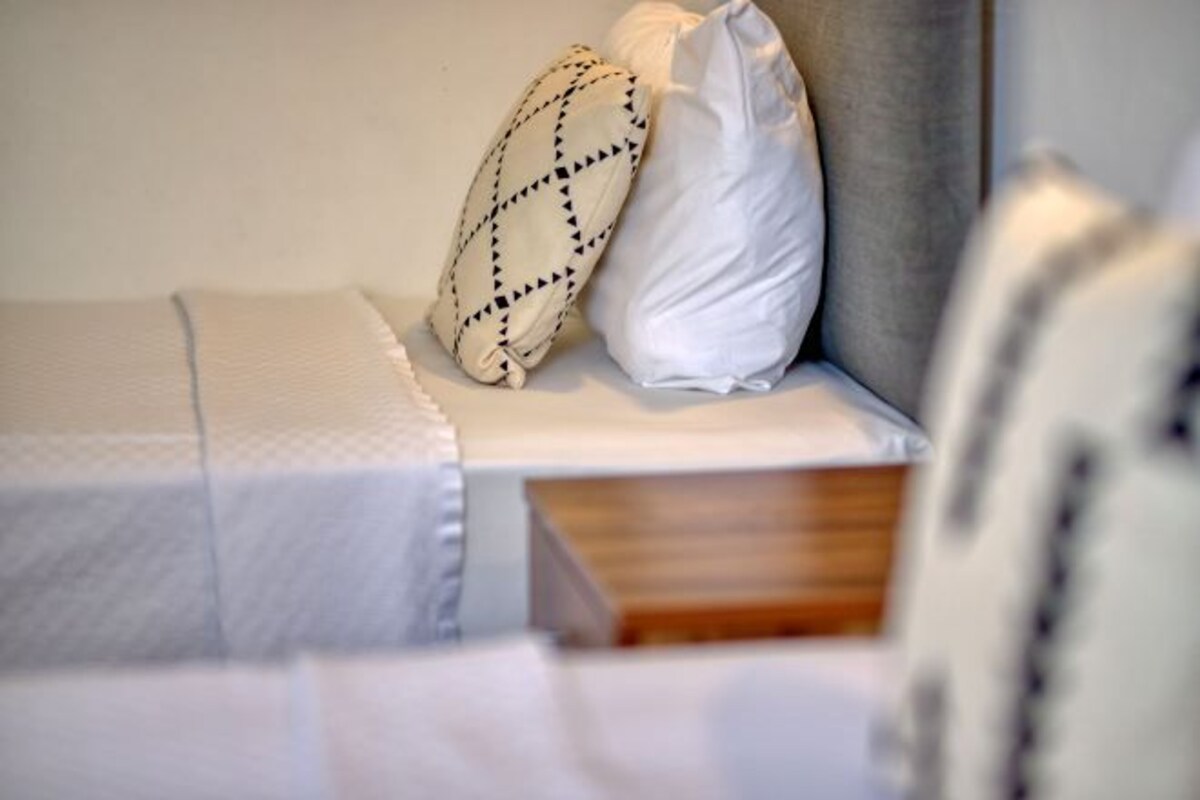 Uma cama bem arrumada com almofada branca e estampada, cabeceira de madeira e parede branca.