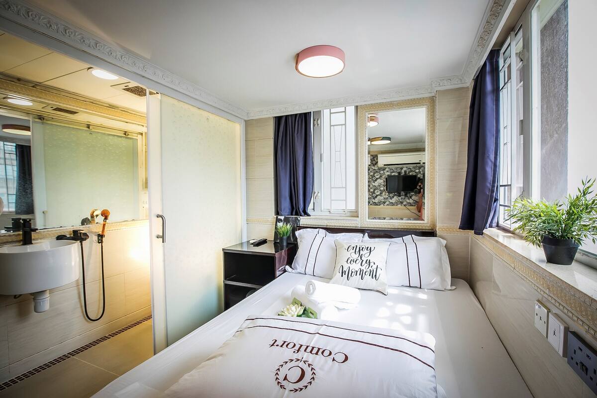 Quarto de hotel com cama, mesa de cabeceira, espelho, janela com cortinas, pia e planta em vaso.