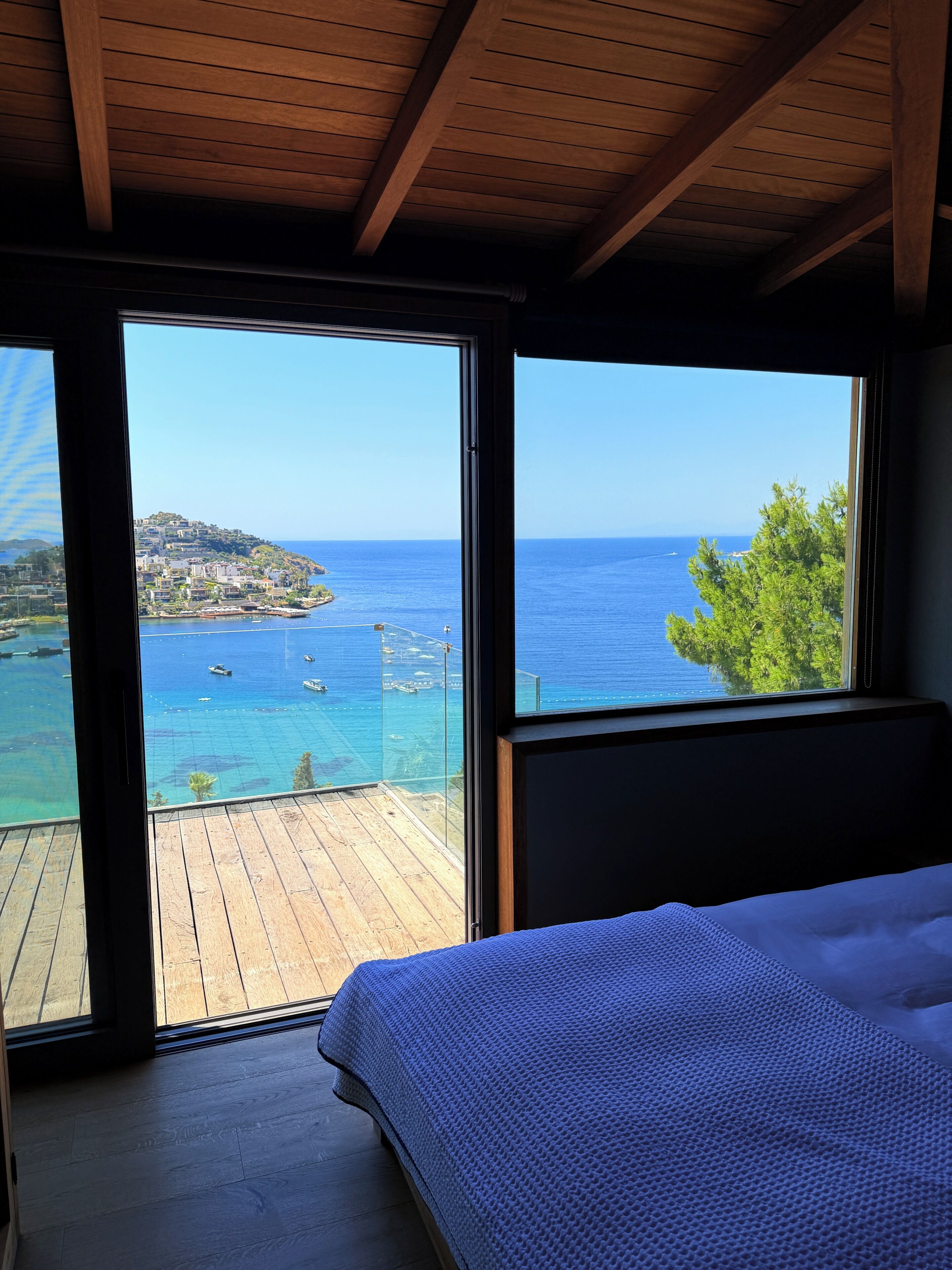 Quarto com uma grande janela com vista para a costa, piso de madeira e uma cama com colcha azul texturizada.