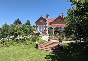 Uma casa de tijolos com telhado vermelho, varanda frontal e um jardim bem cuidado com flores e gramado.