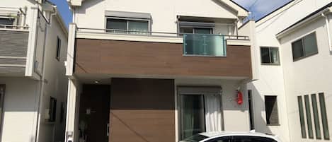 Um edifício residencial de dois andares com fachada branca, uma varanda e um carro branco estacionado na frente.