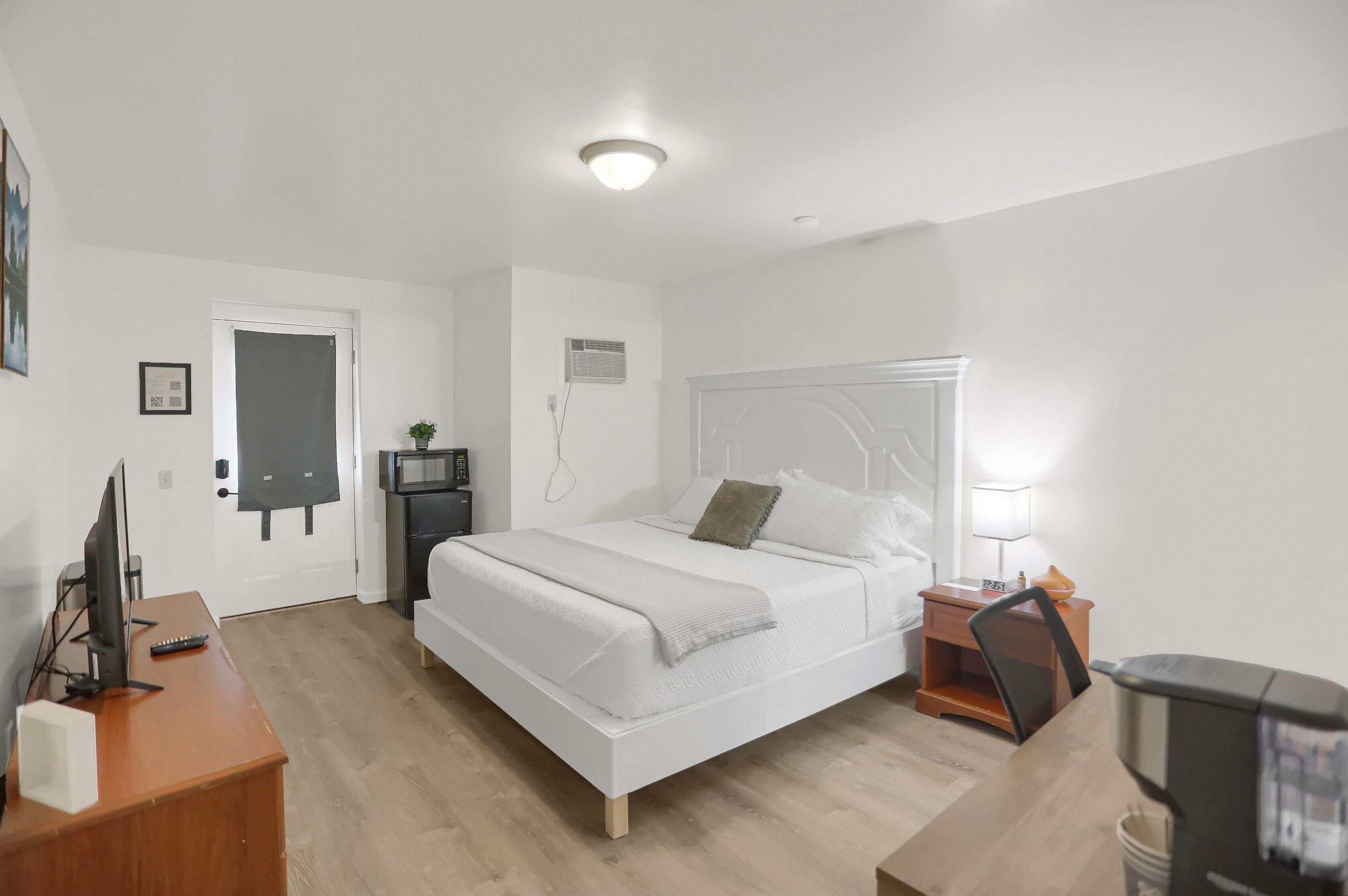 Quarto de hotel moderno com uma cama grande, uma escrivaninha com computador, uma TV e uma mesinha de cabeceira com abajur.