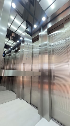 Interior de elevador com paredes metálicas, luminárias no teto e botões de comando.