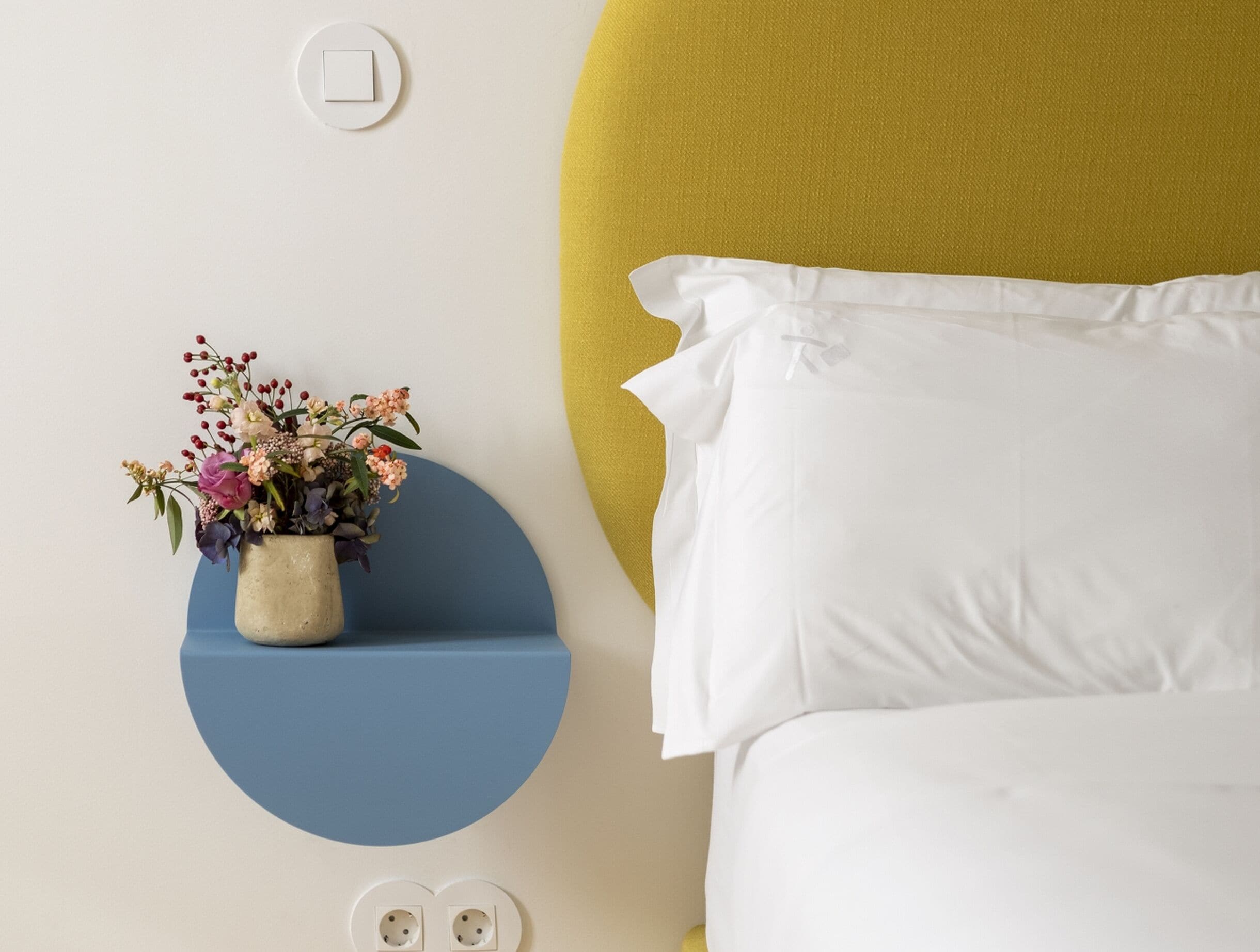 Uma cama com roupa de cama branca, cabeceira amarela, prateleira circular azul com arranjo floral e parede branca com interruptor de luz e tomadas.