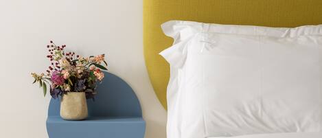 Uma cama com roupa de cama branca, cabeceira amarela, prateleira circular azul com arranjo floral e parede branca com interruptor de luz e tomadas.