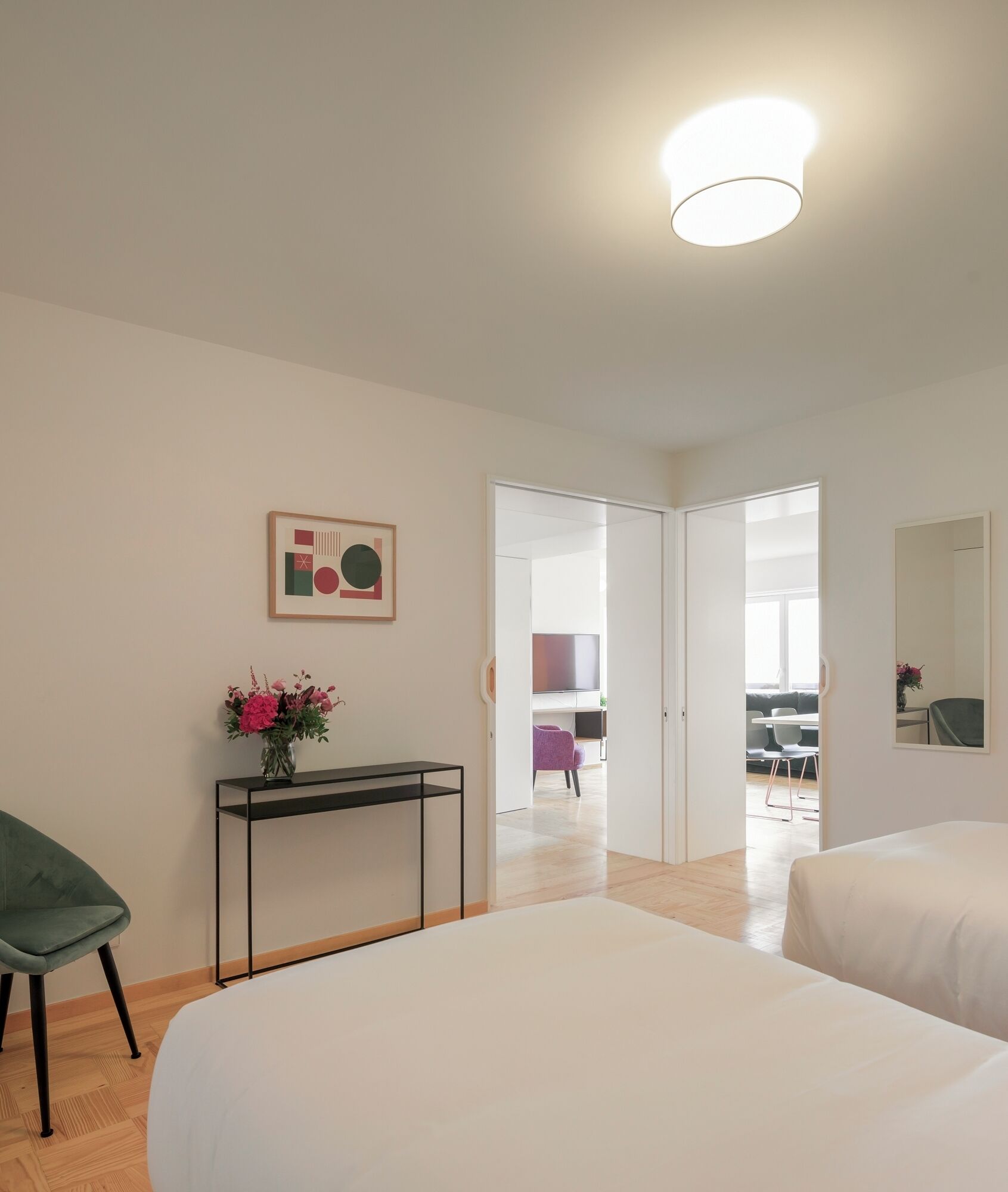 Quarto de hotel com duas camas, uma escrivaninha com um vaso de flores, um quadro na parede e vista para a área de estar adjacente.