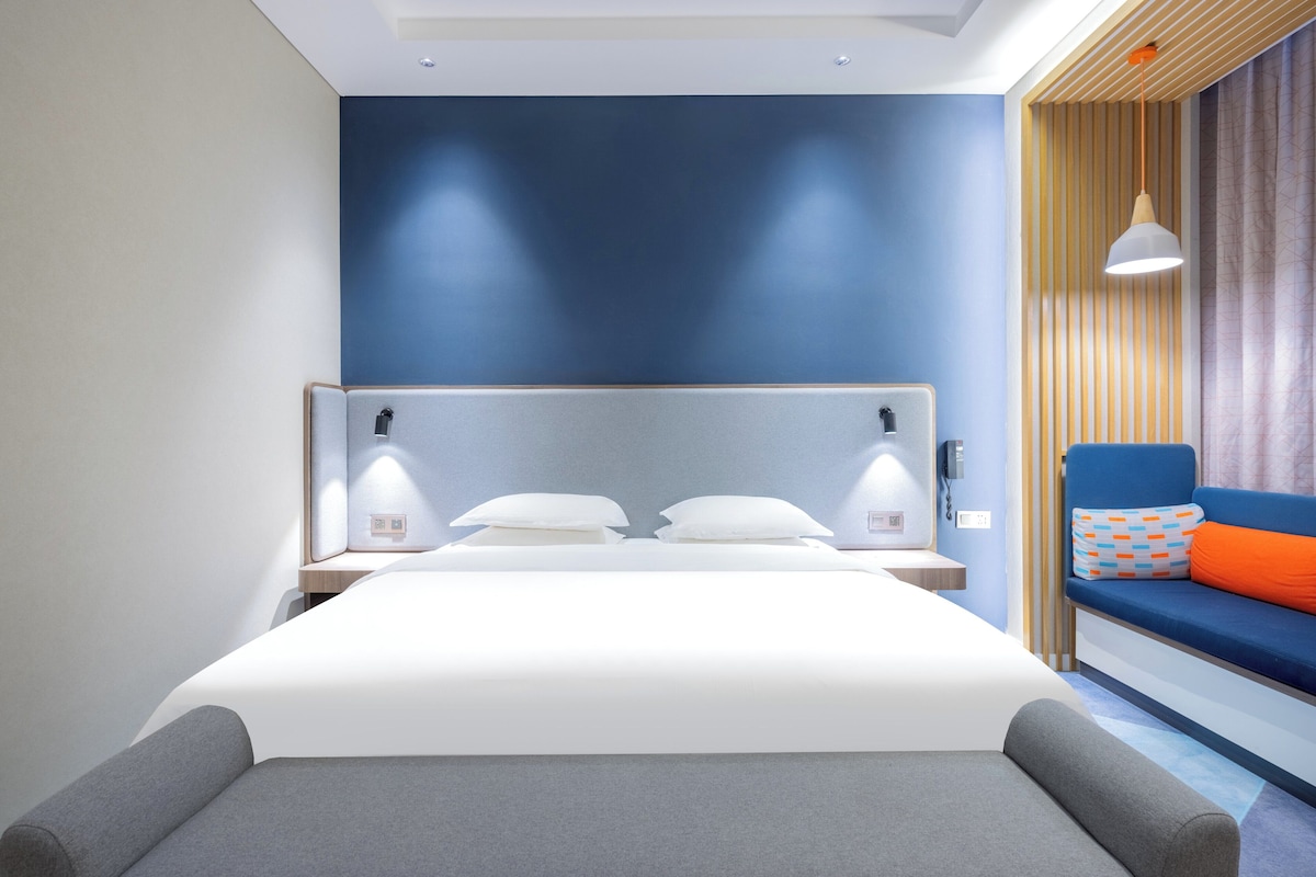 Quarto de hotel moderno com uma cama grande, luminárias de cabeceira, uma parede azul de destaque e um espaço de estar com sofá e almofadas.