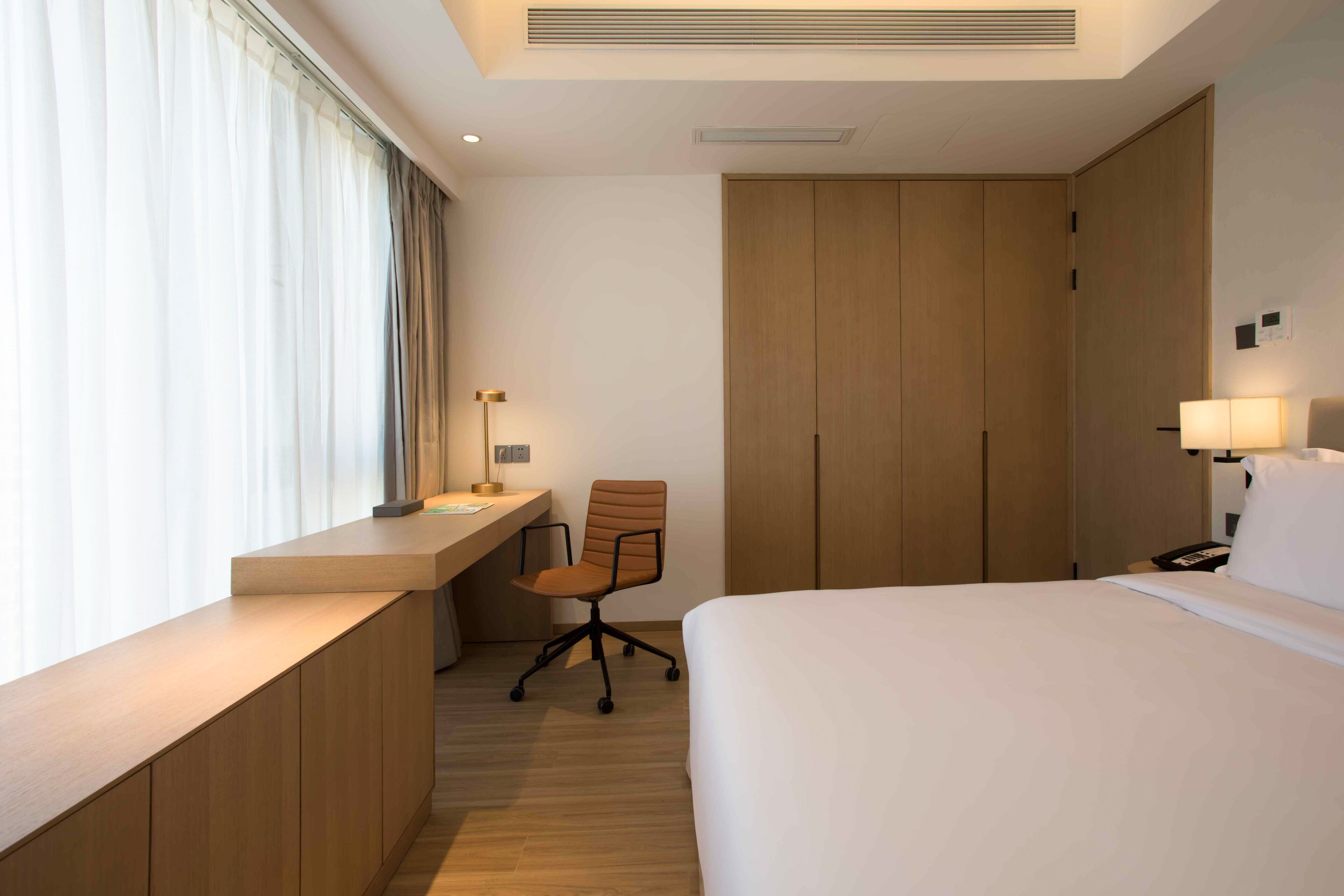 Quarto de hotel com cama, escrivaninha, cadeira, guarda-roupa e janela com cortinas.