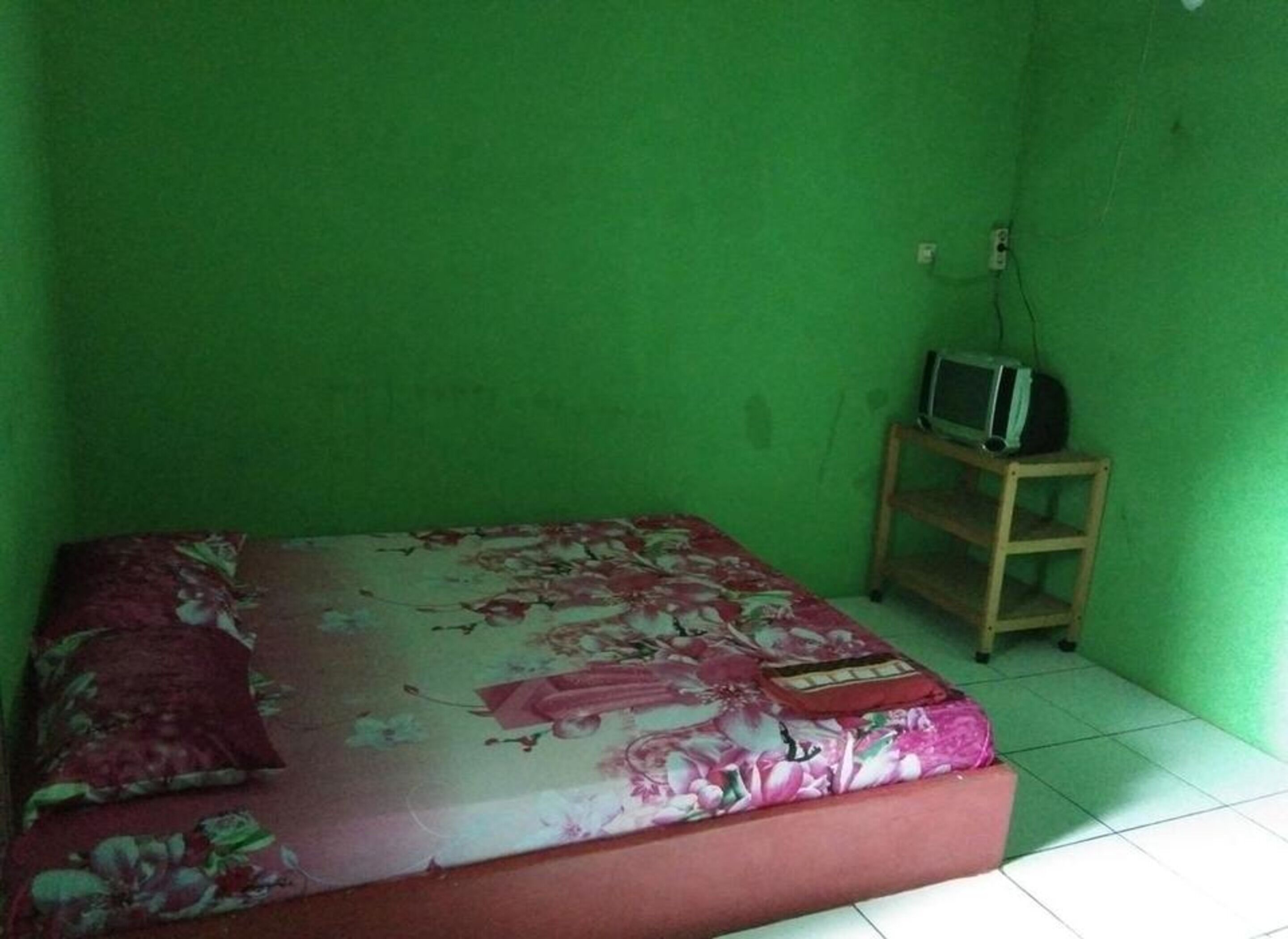 Quarto com paredes verdes, cama com roupa de cama floral e uma pequena prateleira de madeira com uma televisão.