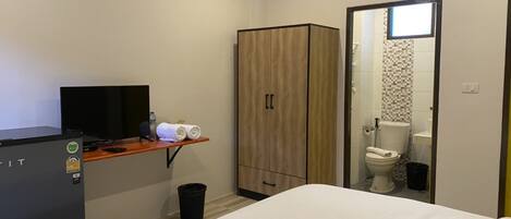 Quarto de hotel com uma cama, televisão, guarda-roupa e banheiro visível por uma porta aberta.