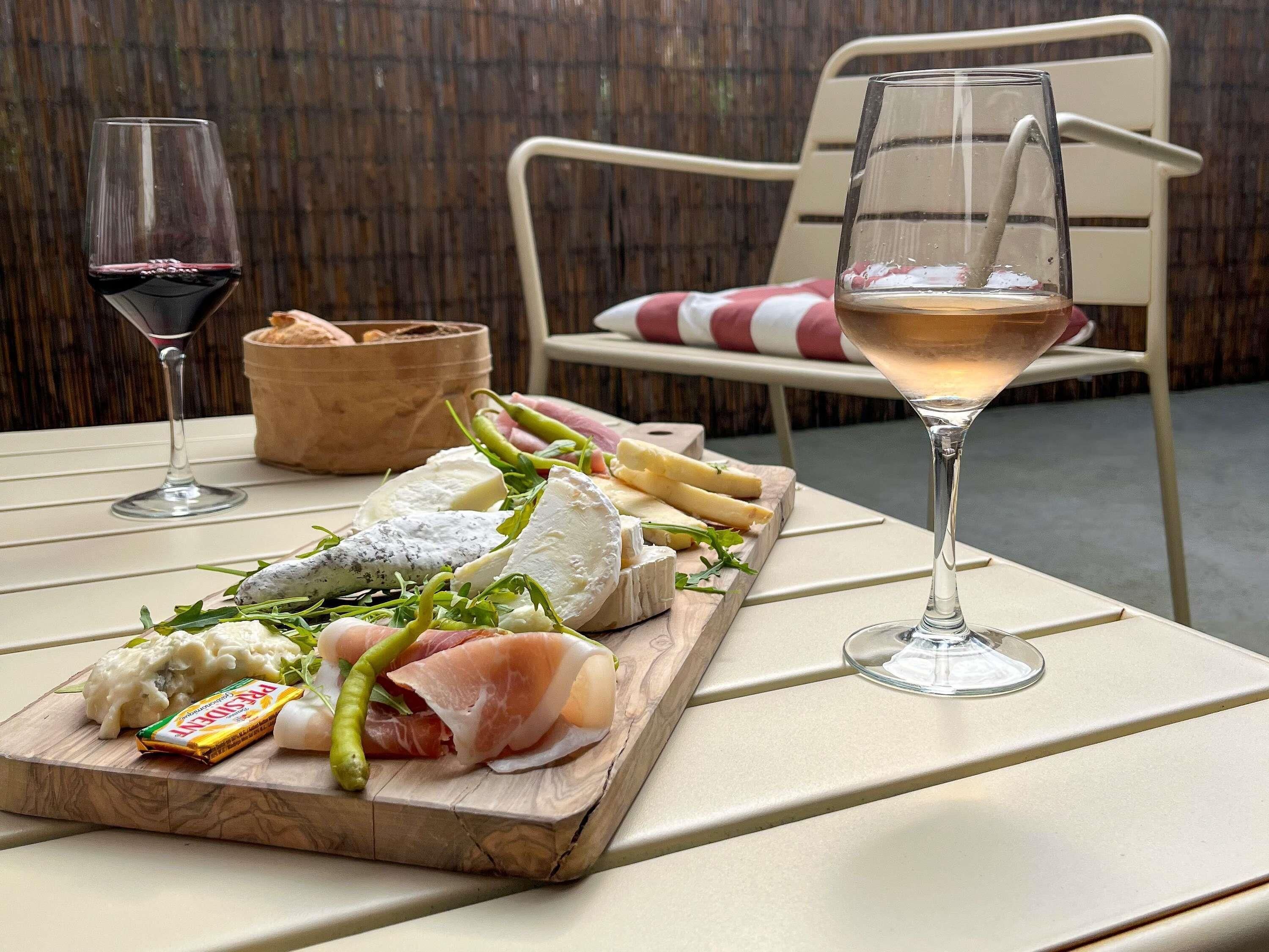 Uma mesa com uma tábua de queijos, dois copos de vinho e uma cadeira.