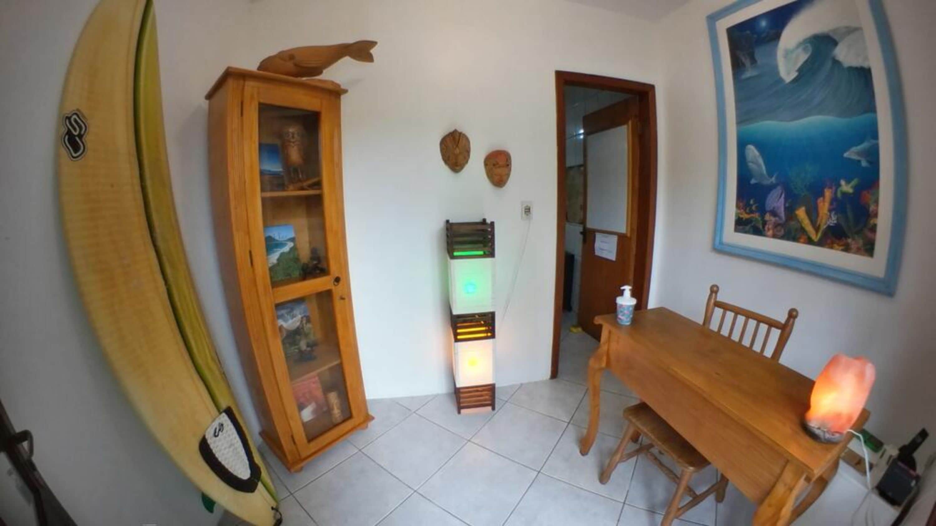 Quarto com armário de madeira, uma prancha de surf, mesa com cadeira, aquecedor fixado na parede e um quadro emoldurado.