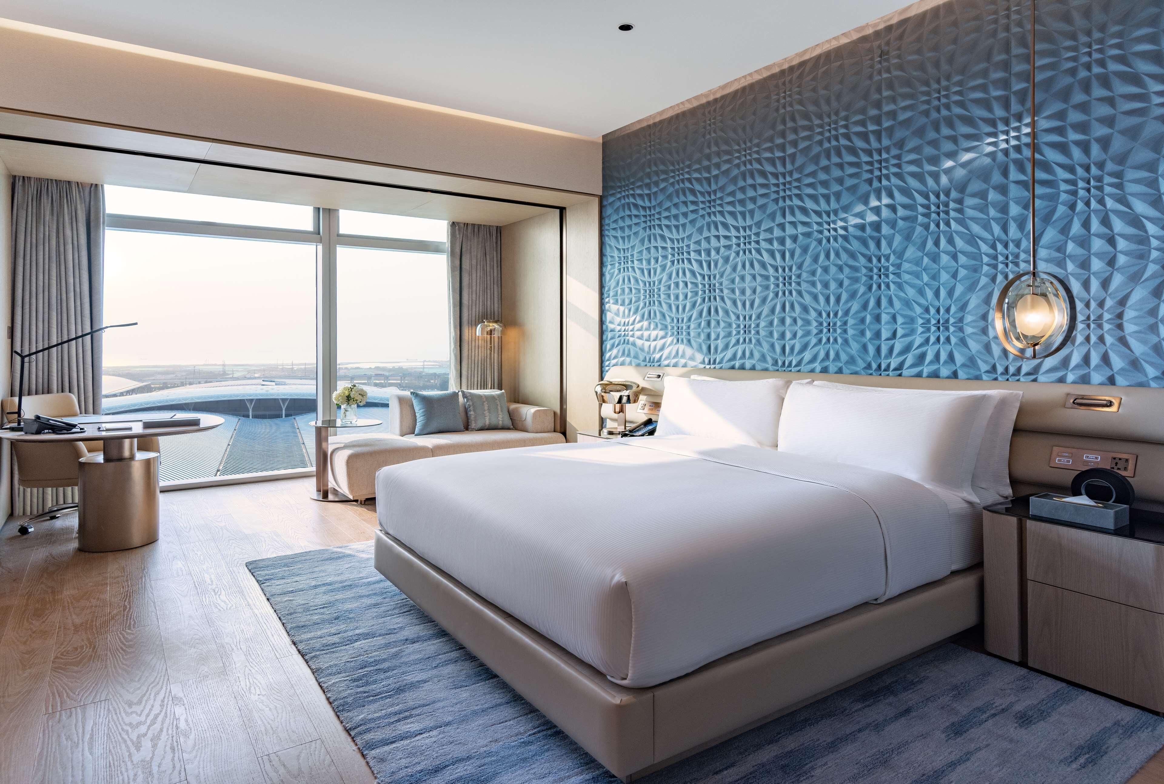 Quarto de hotel moderno com uma cama grande, uma escrivaninha e vista para a paisagem urbana através de janelas amplas.
