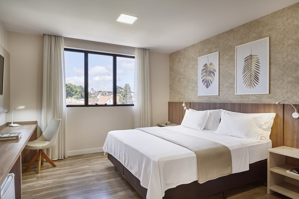 Quarto de hotel moderno com uma cama grande, uma escrivaninha e uma cadeira. Há uma janela com vista para edifícios e árvores.