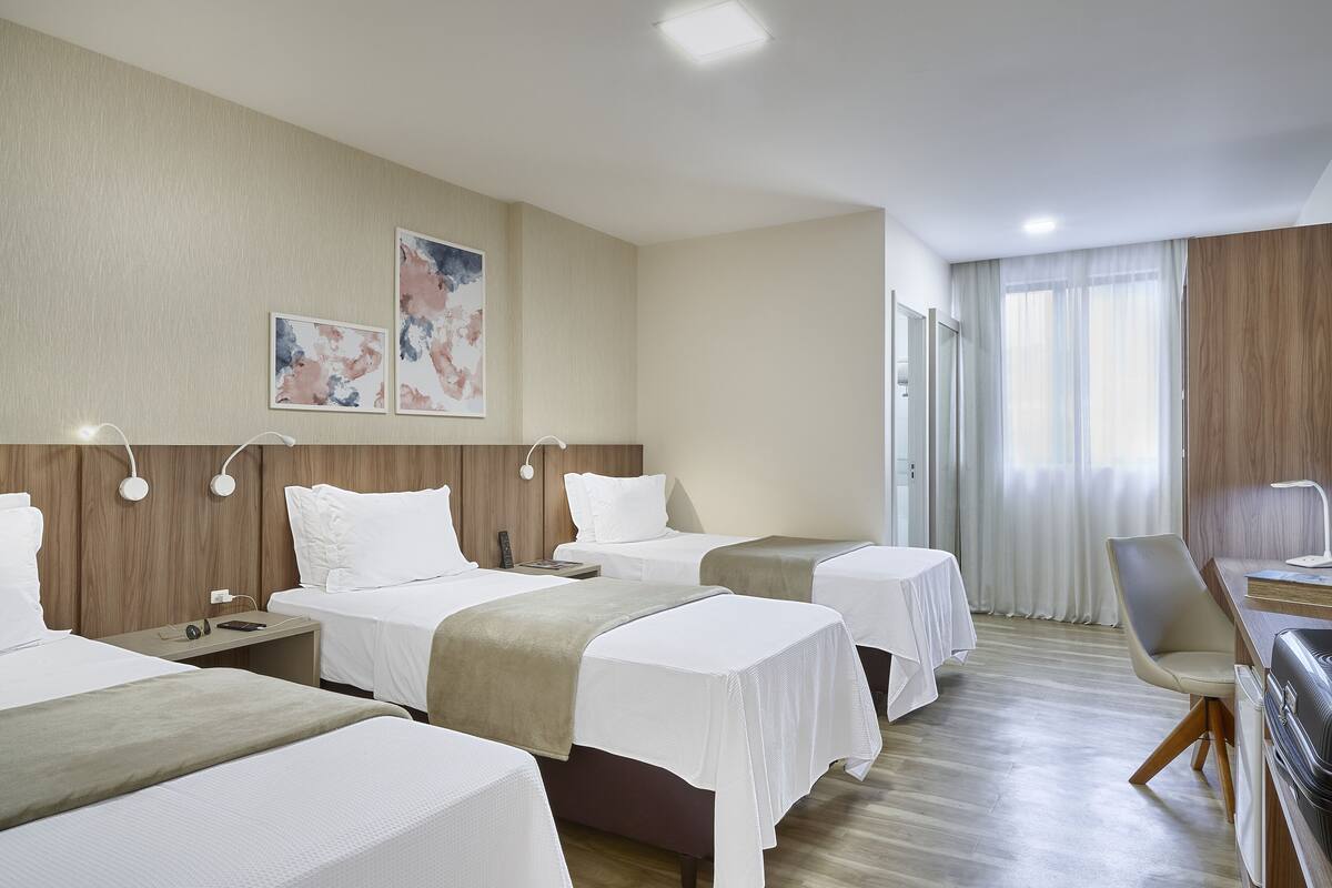 Quarto de hotel com três camas, uma mesa, uma cadeira e uma janela grande com cortinas.
