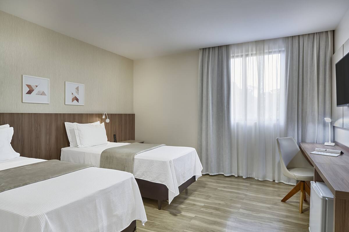 Quarto de hotel com duas camas, uma mesa, uma cadeira e uma televisão.
