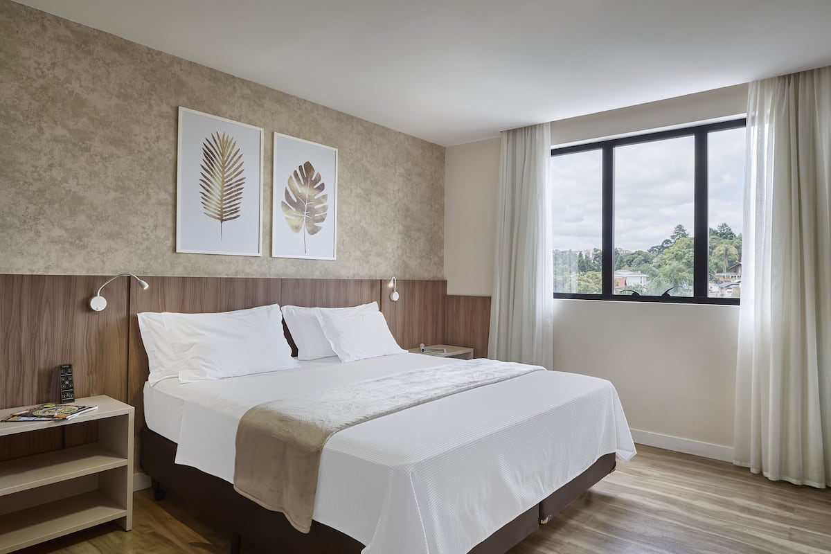 Quarto de hotel com uma cama grande, uma mesa de cabeceira, luminária na parede e uma janela com cortinas.