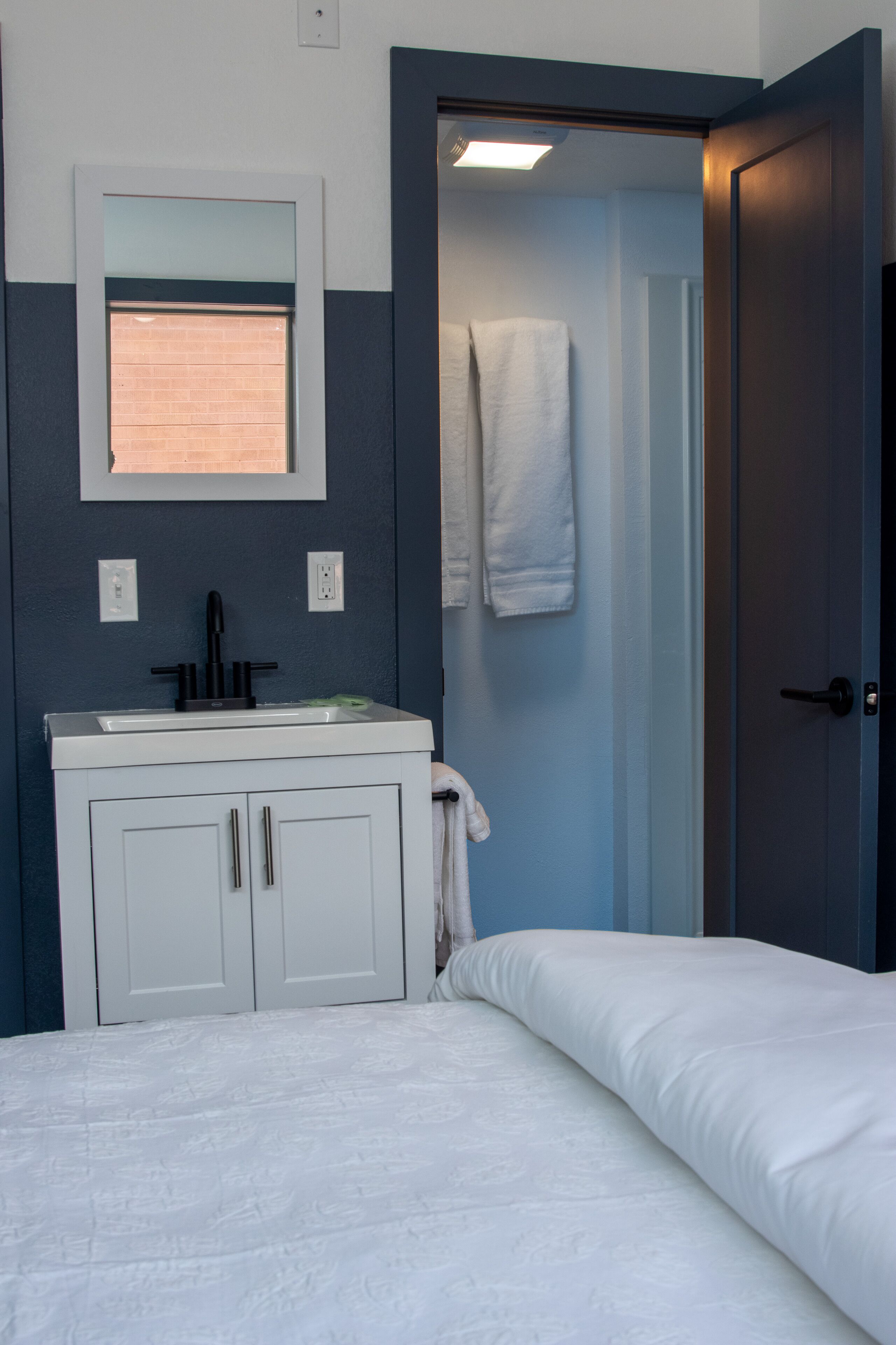 Quarto com uma cama branca, um armário branco com uma torneira preta e uma porta que dá para um banheiro com uma toalha pendurada na parede.