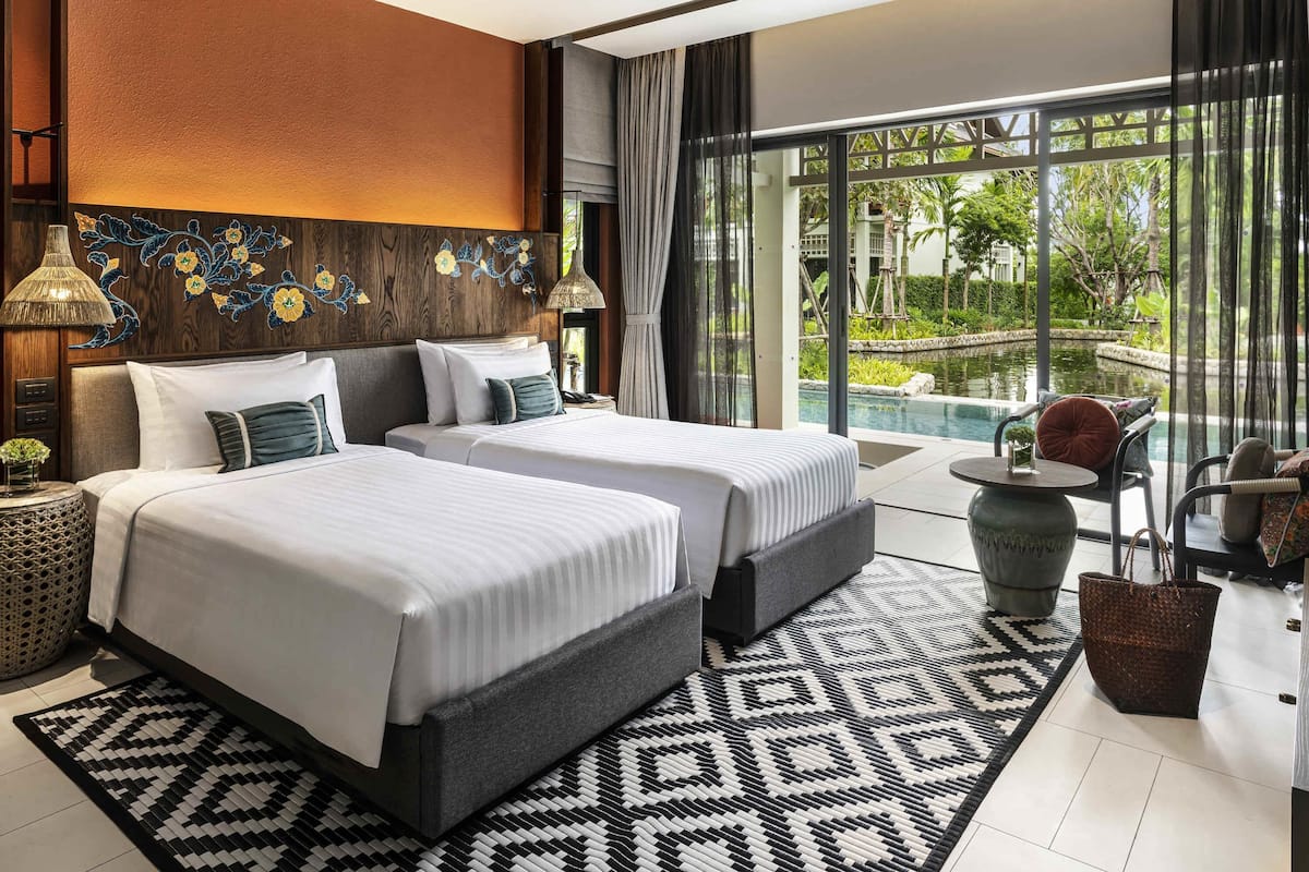 Quarto de hotel com duas camas, uma janela grande com vista para uma piscina, um tapete tecido e um painel decorativo na parede.