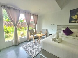 Quarto de hotel com cama, uma mesinha, duas cadeiras, janela com cortinas e vista para o verde lá fora.