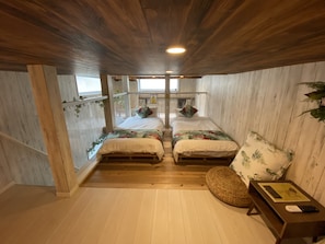 Quarto com duas camas de madeira, uma mesinha com uma planta e uma janela com vista.
