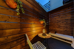 Um deck de madeira com um banco, uma mesa pequena com luminária e vasos suspensos.