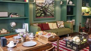 Uma sala de estar aconchegante com um sofá verde, uma mesinha de centro com lanches, uma estante de livros e um quadro de uma árvore.