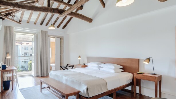 Quarto com cama de madeira, mesas de cabeceira, um banco e vista para o exterior.