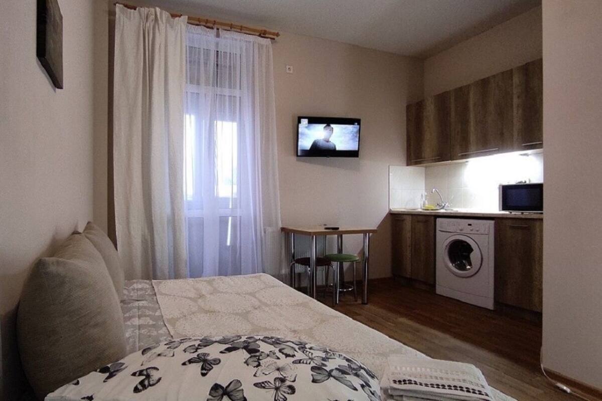 Quarto pequeno e bem iluminado com cama, televisão, máquina de lavar e cozinha compacta.