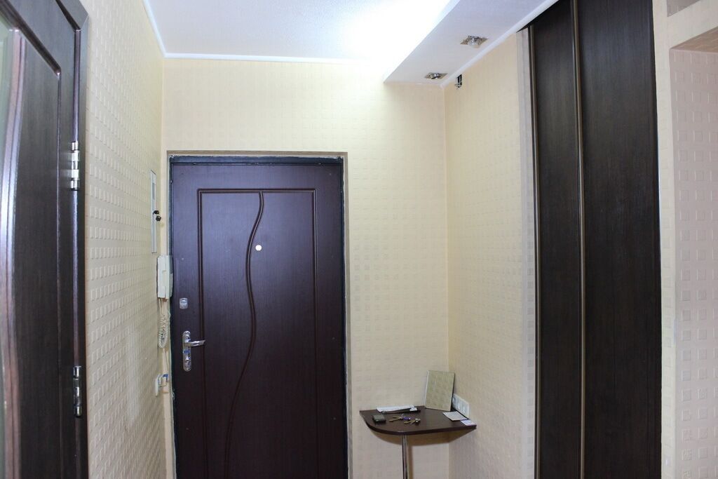 Um corredor estreito com uma porta de madeira escura, uma pequena mesa com uma cadeira e um interruptor de luz na parede.