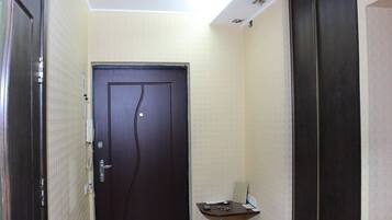 Um corredor estreito com uma porta de madeira escura, uma pequena mesa com uma cadeira e um interruptor de luz na parede.