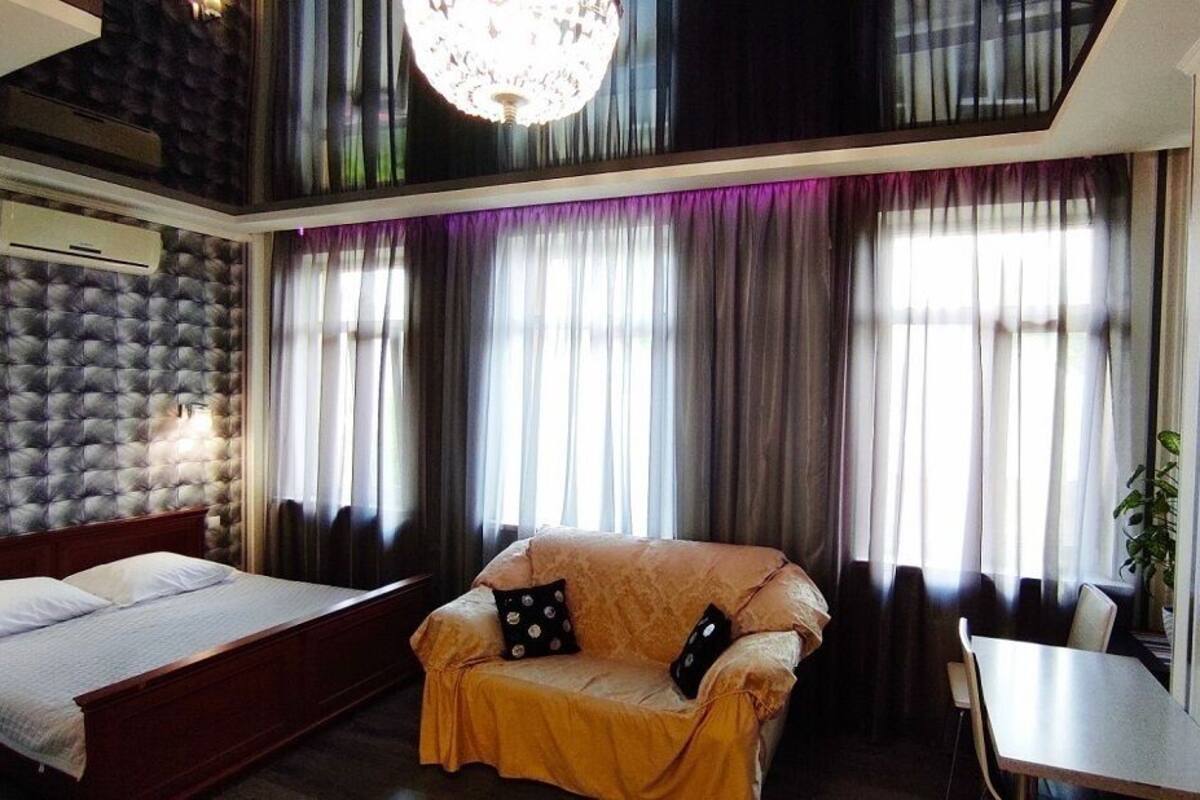 Quarto de hotel com cama, sofá, televisão e uma janela grande com cortinas.