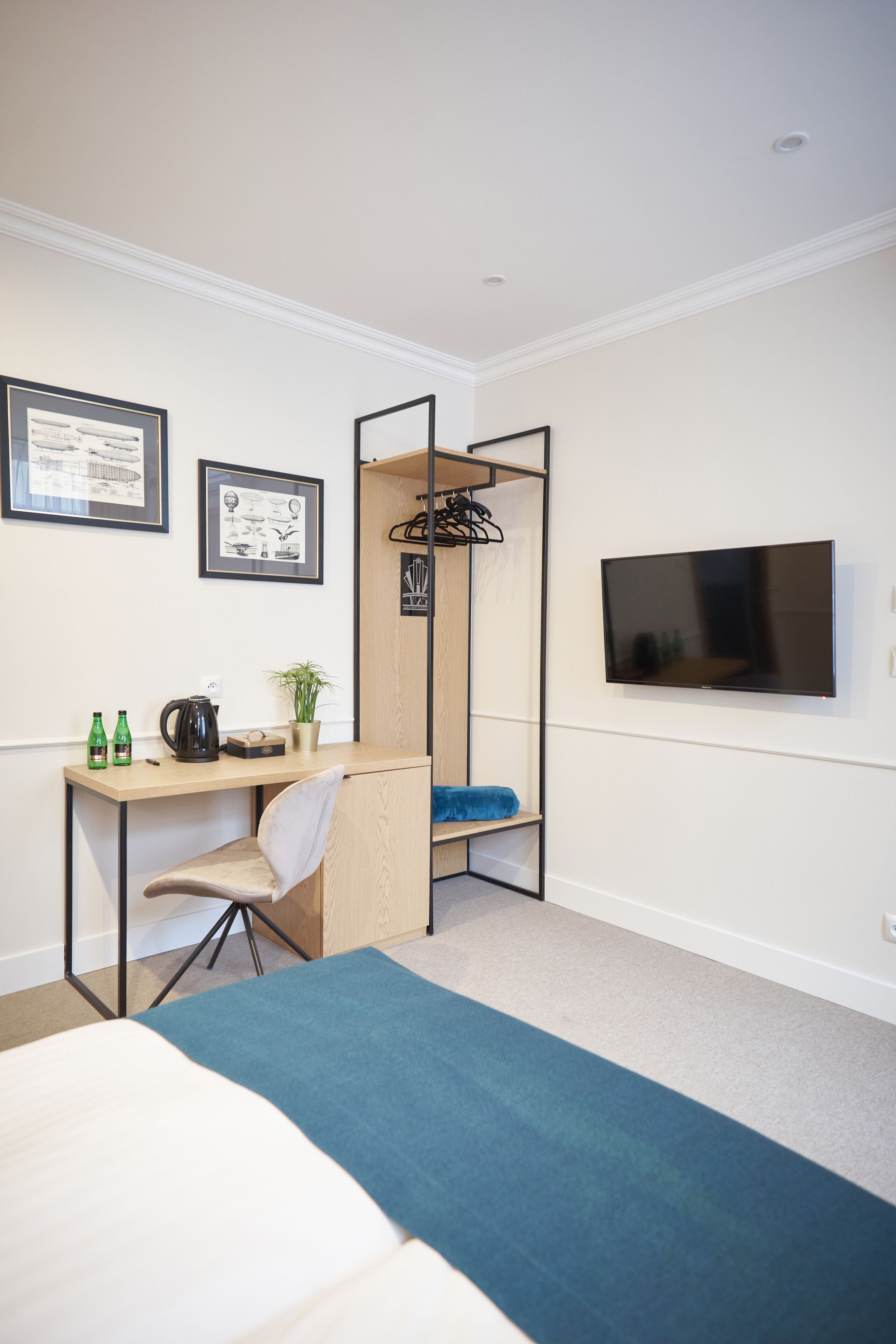 Quarto de hotel compacto com uma cama, uma mesa com cadeira, uma TV fixada na parede e uma prateleira pequena com uma planta e uma chaleira.