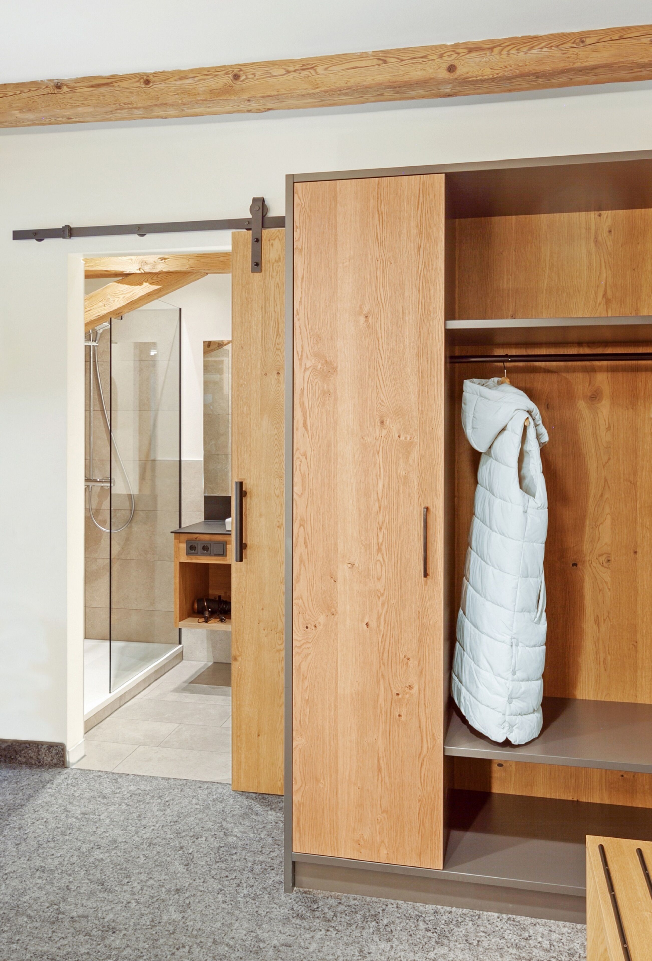Um guarda-roupa de madeira com porta de correr, um cobertor branco acolchoado pendurado no interior, e uma área de chuveiro visível através de uma porta de vidro.