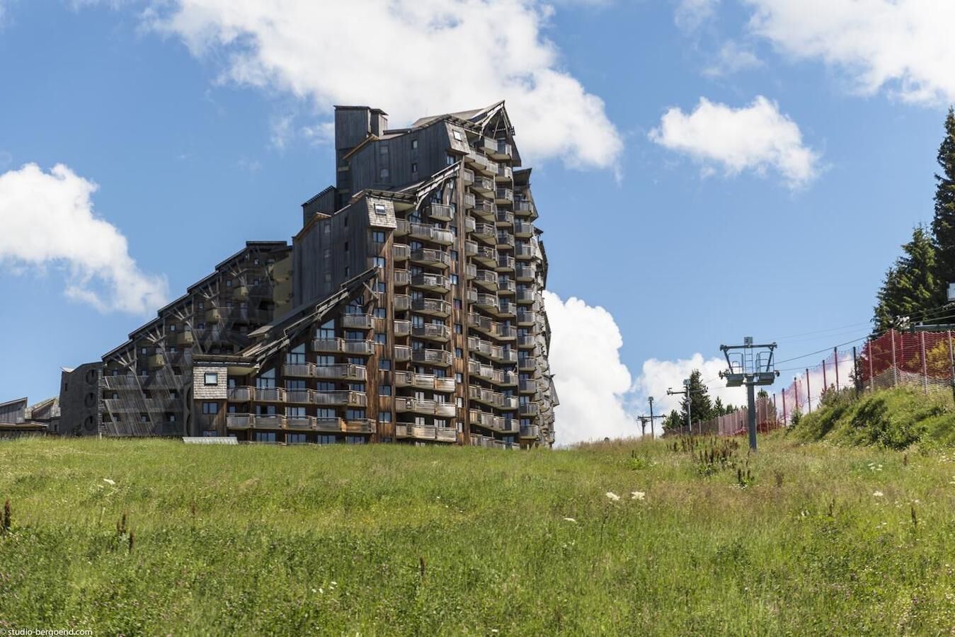 Um edifício moderno de vários andares com varandas, situado numa área ajardinada sob um céu claro.