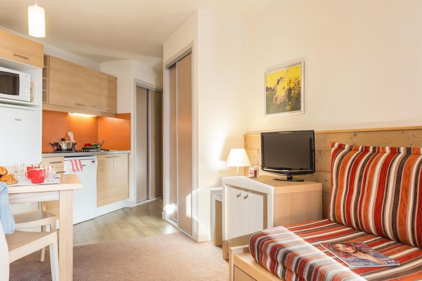 Quarto de hotel pequeno com cozinha compacta, uma cama com roupa de cama listrada, televisão e uma pequena mesa de cabeceira com abajur.