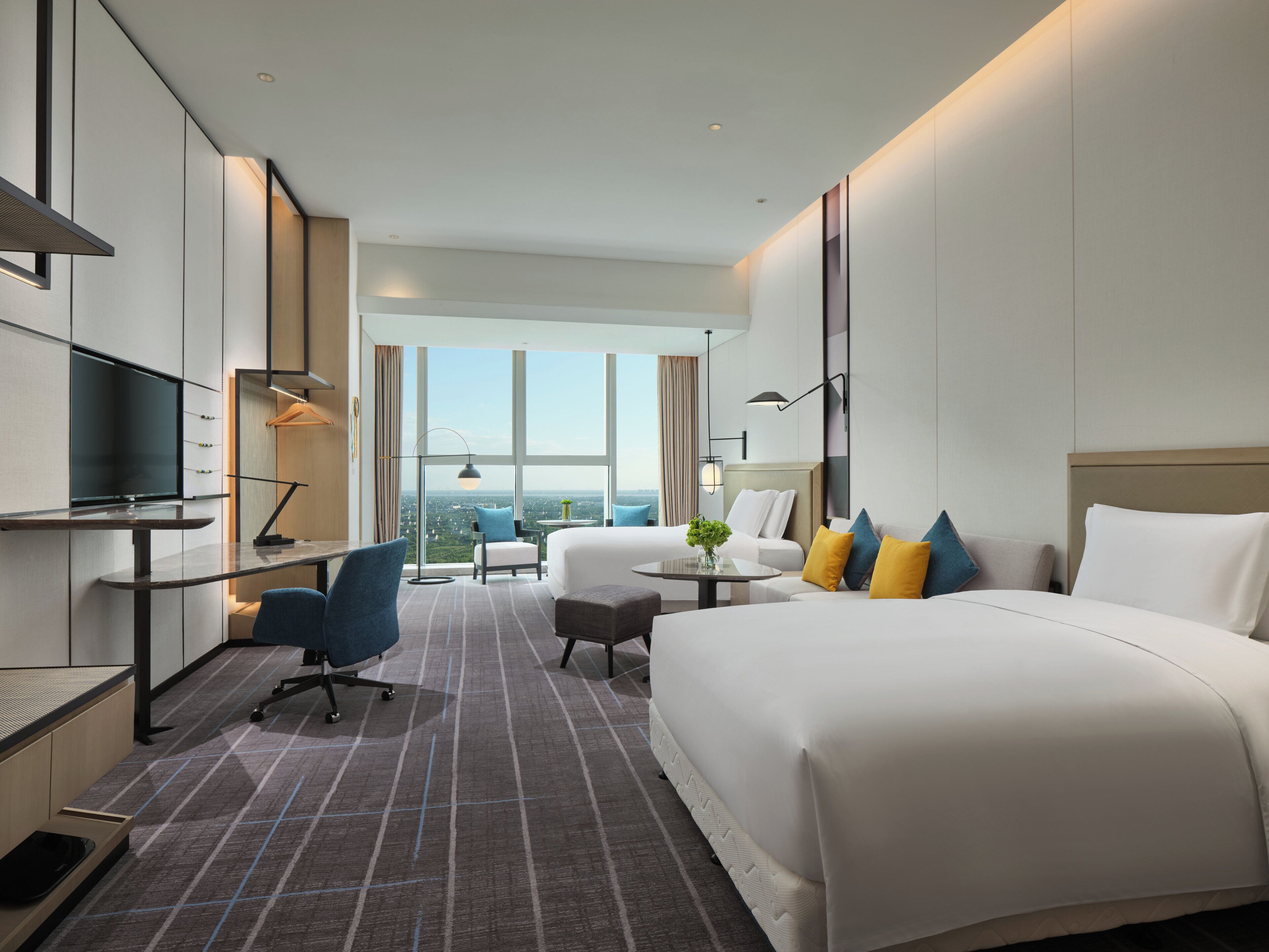 Quarto de hotel moderno com uma cama grande, uma escrivaninha com televisão de tela plana, uma poltrona confortável e vista para a cidade por grandes janelas.