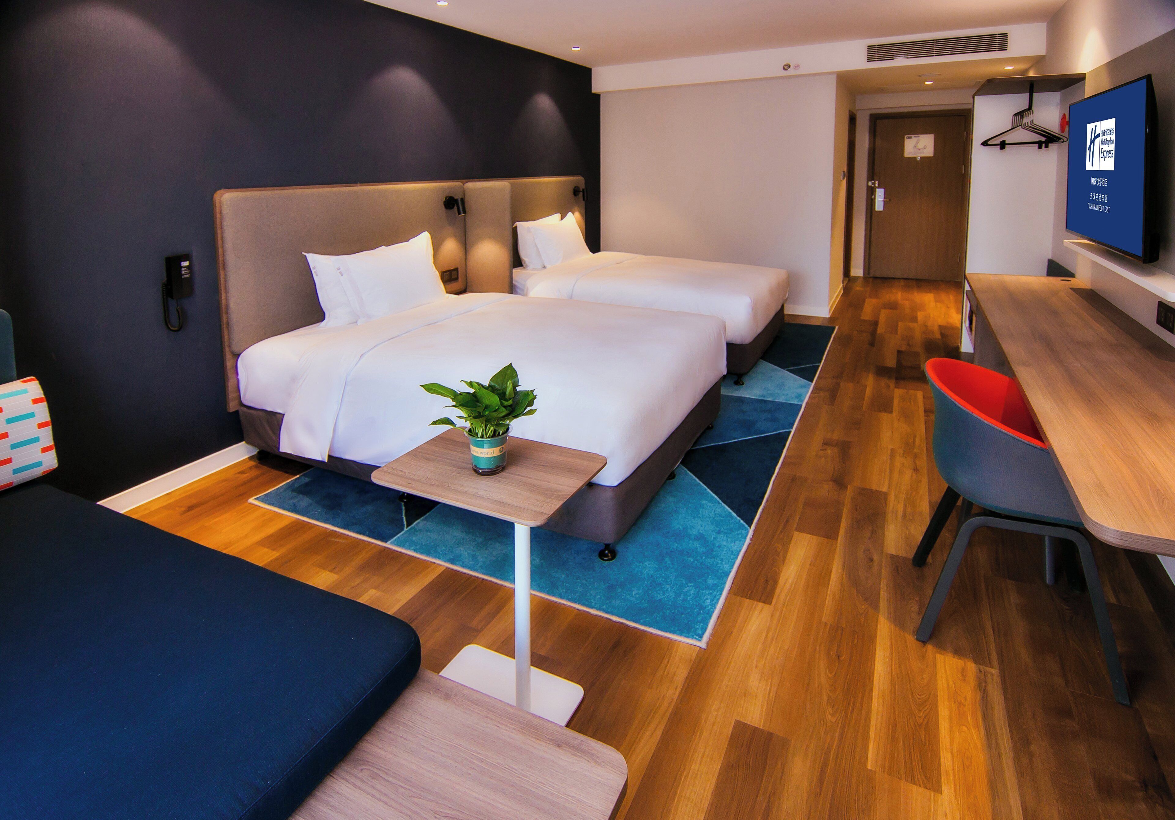 Quarto de hotel com duas camas, uma mesinha com uma planta, uma escrivaninha com uma cadeira e uma televisão na parede.
