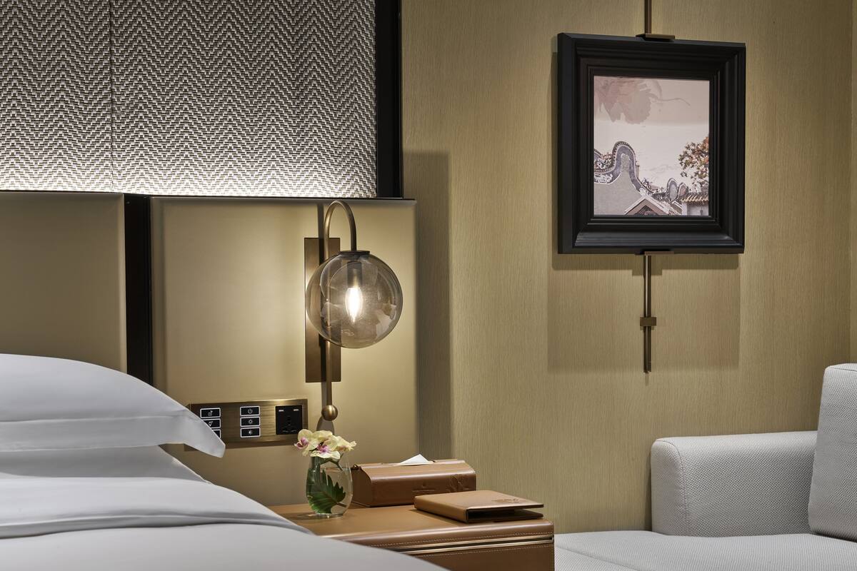 Quarto de hotel com cama, mesa de cabeceira com um vaso de flores, quadro na parede e uma luminária moderna.