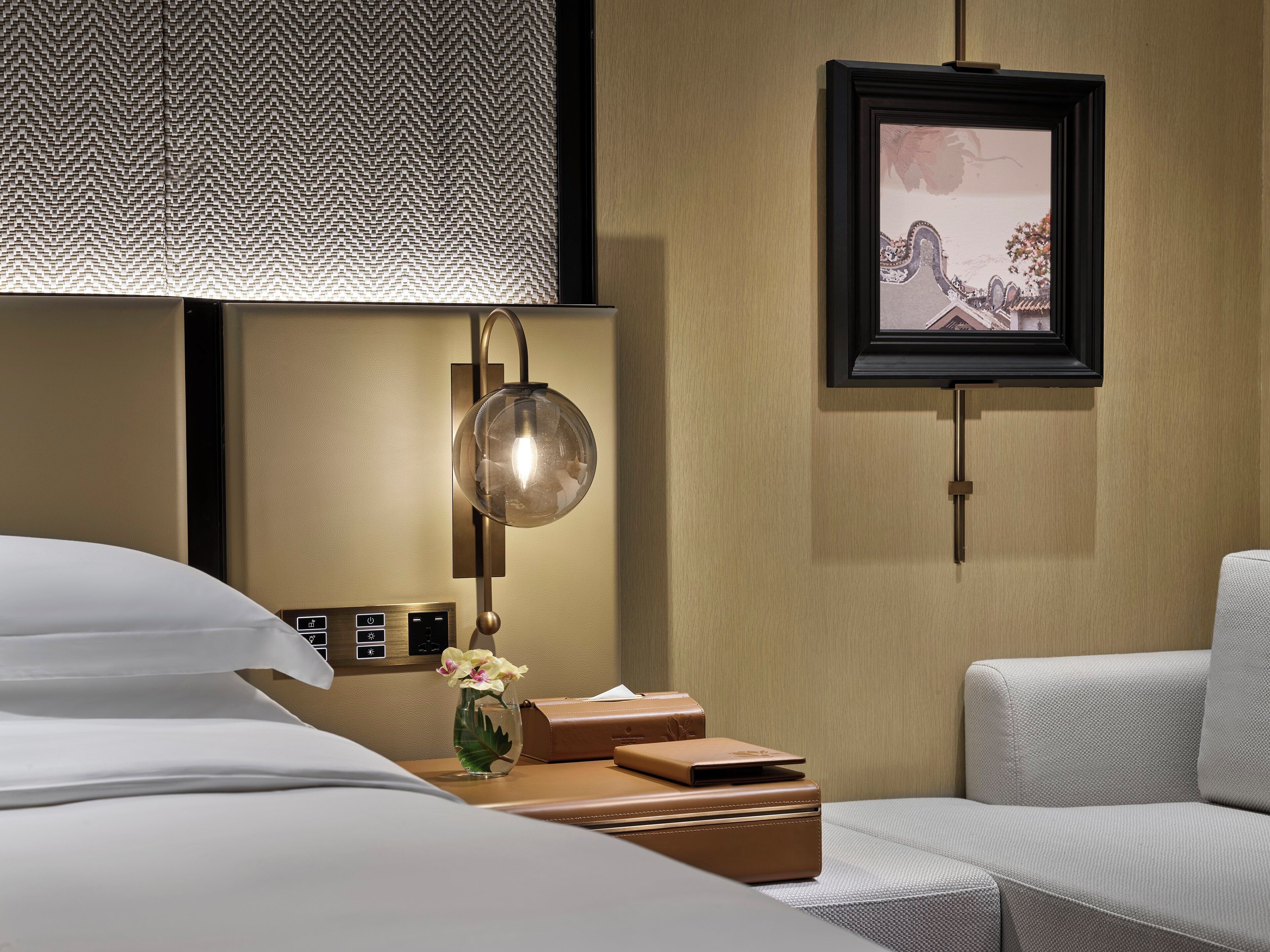 Quarto de hotel com cama, mesa de cabeceira com um vaso de flores, quadro na parede e uma luminária moderna.